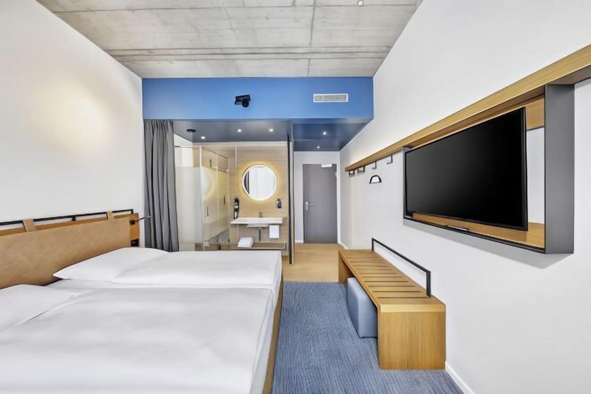 Quarto de hotel com duas camas, televisão, banheiro com espelho e uma escrivaninha.