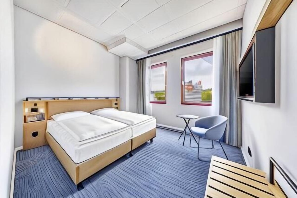 Quarto de hotel com cama de casal, uma mesinha, uma cadeira, televisão e janelas amplas.