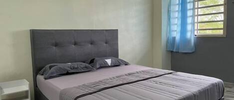 Cama com cabeceira cinza e almofadas, mesa de cabeceira e janela com cortinas azuis.