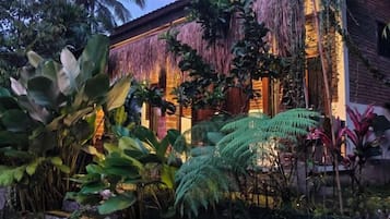 Um jardim tropical com um edifício de telhado de palha, vegetação exuberante e um gramado bem cuidado.