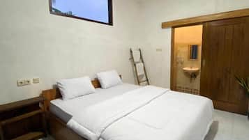 Quarto com uma cama grande, mesa de cabeceira de madeira, janela, porta de madeira e banheiro pequeno com pia e espelho.