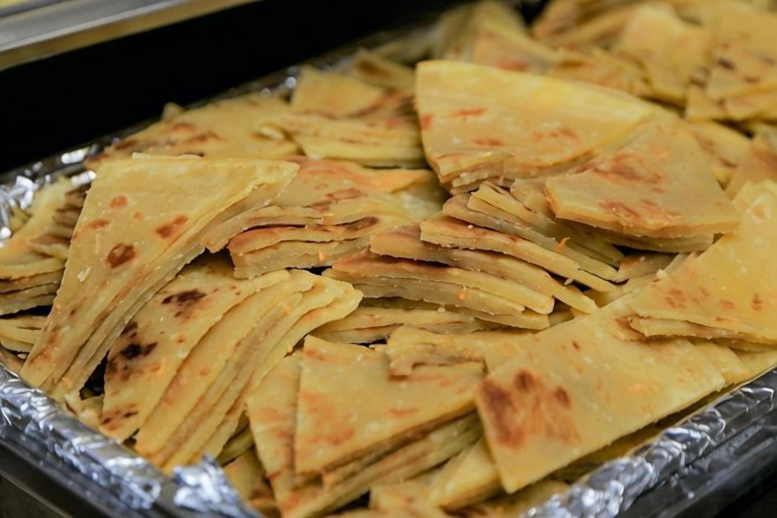 Uma bandeja com porções triangulares de comida, possivelmente um tipo de pão achatado, dispostas sobre uma superfície forrada com papel alumínio.