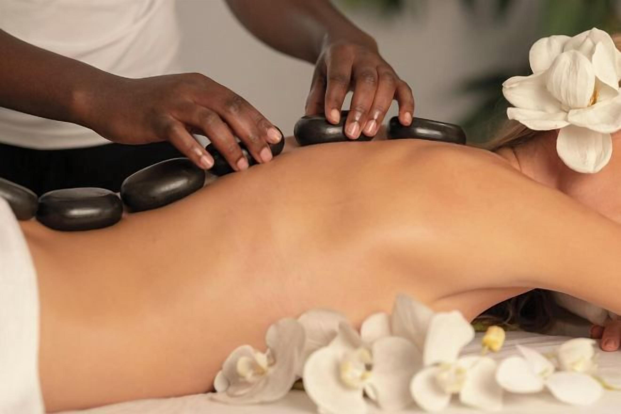 Uma pessoa recebendo massagem com pedras e flores brancas sobre a mesa.