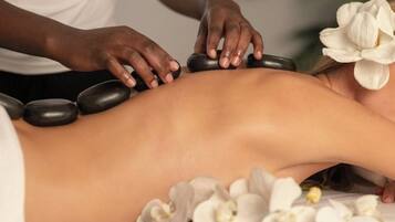 Uma pessoa recebendo massagem com pedras e flores brancas sobre a mesa.