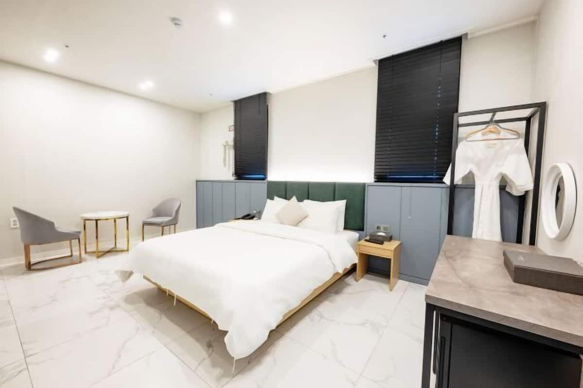 Quarto de hotel moderno com uma cama grande, mesa de cabeceira, uma cadeira, uma mesa redonda pequena e um espelho.