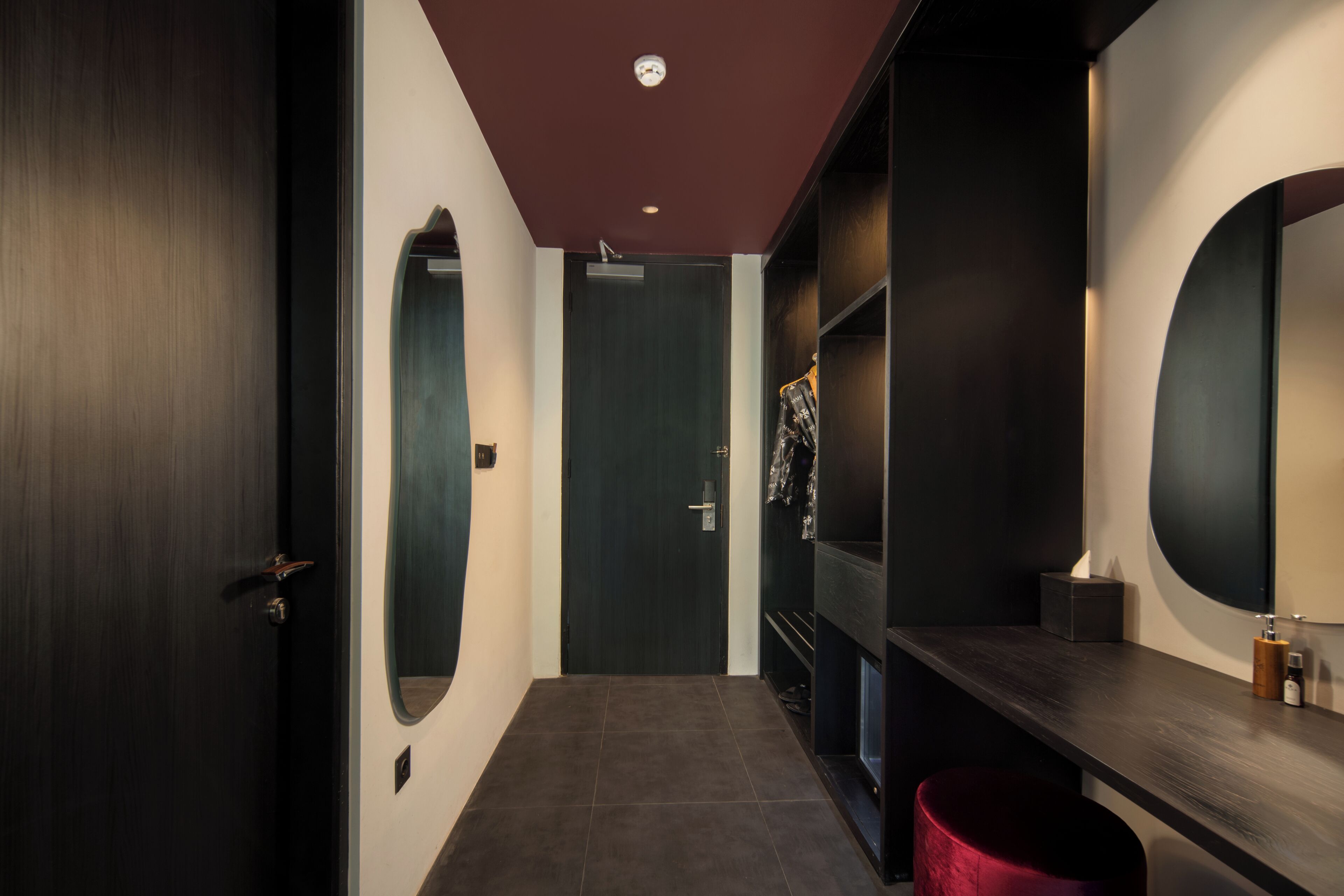 Um corredor com uma porta escura, um espelho, uma prateleira com objetos e um banquinho vermelho.