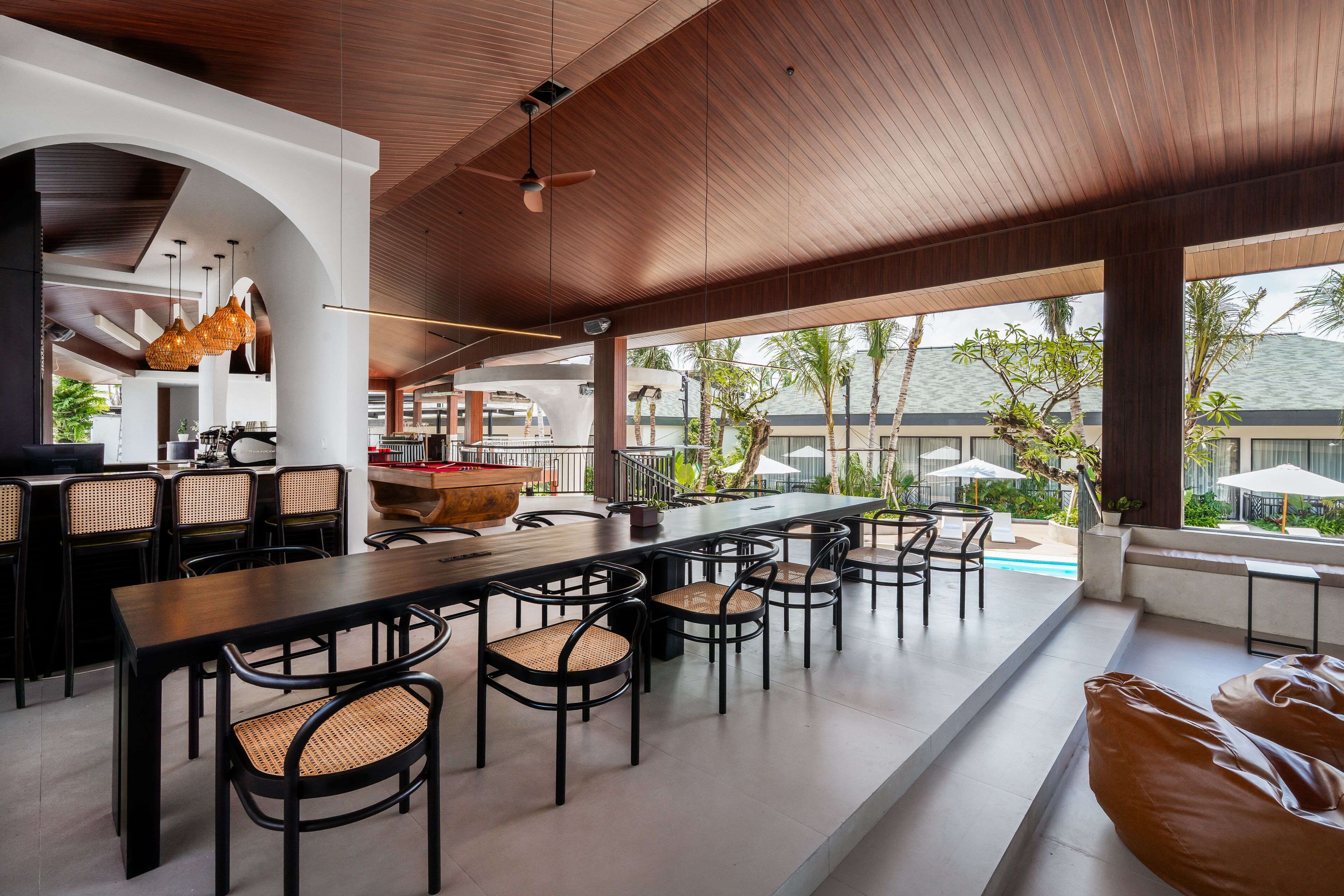 Uma área de refeições ao ar livre ampla, com uma mesa de madeira comprida, cadeiras pretas e vista para a piscina e palmeiras.