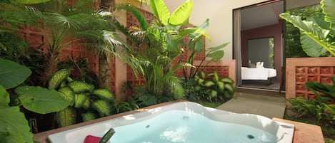 Uma banheira de hidromassagem rodeada por vegetação tropical exuberante, cerca de madeira e vislumbre de um quarto com toalha branca.