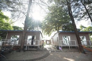 Duas cabanas com varandas externas, rodeadas por árvores, e uma trilha de terra que leva a um edifício.