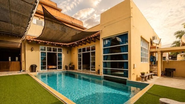 Uma piscina com borda em azulejo, rodeada por gramado e um edifício com janelas amplas e um alpendre coberto.