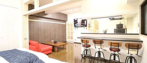 Quarto de hotel moderno com uma cama grande, um sofá vermelho, uma área de cozinha com televisão e um bar com banquetas.