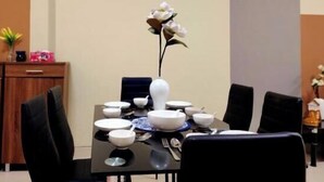 Uma área de refeições com uma mesa posta para quatro pessoas, um vaso com flores e um armário com um vaso e uma caixa.