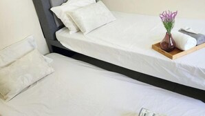Uma cama de casal com roupa de cama branca, cabeceira cinza, um vaso com flores e uma bandeja com um travesseiro e uma toalha.