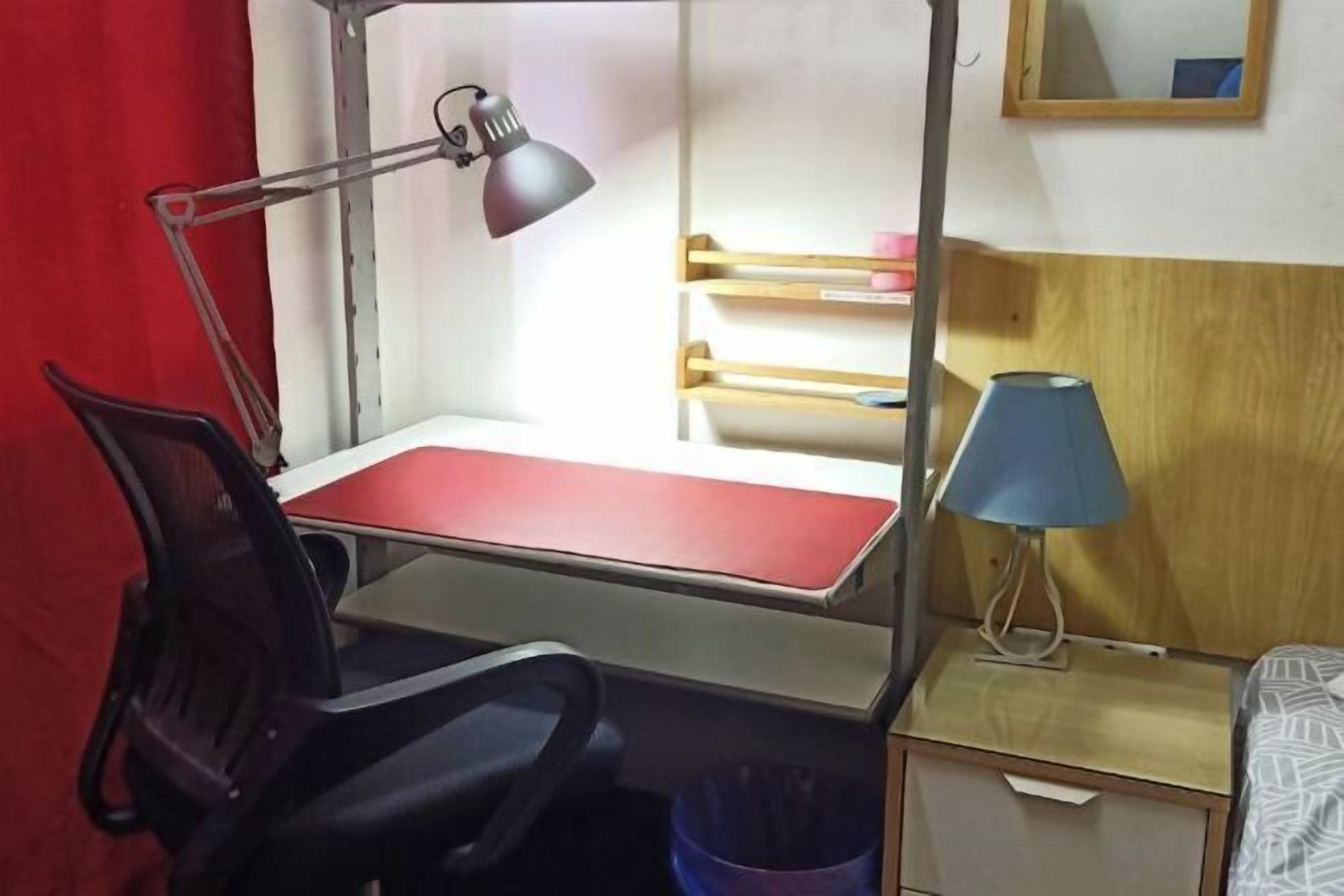 Quarto compacto com escrivaninha, cadeira e cama. Há uma luminária de mesa, uma luminária de cabeceira e uma pequena lixeira.