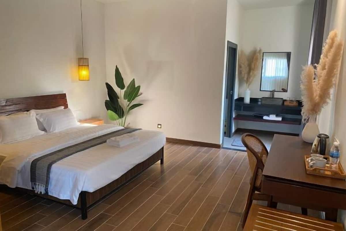 Quarto de hotel com cama, mesa de cabeceira, luminária, planta em vaso e um espaço de refeições com mesa e cadeiras.