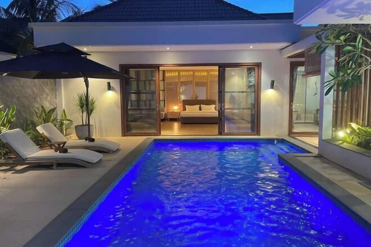 Uma área moderna à beira da piscina com quarto em vidro, espreguiçadeiras e interior bem iluminado.
