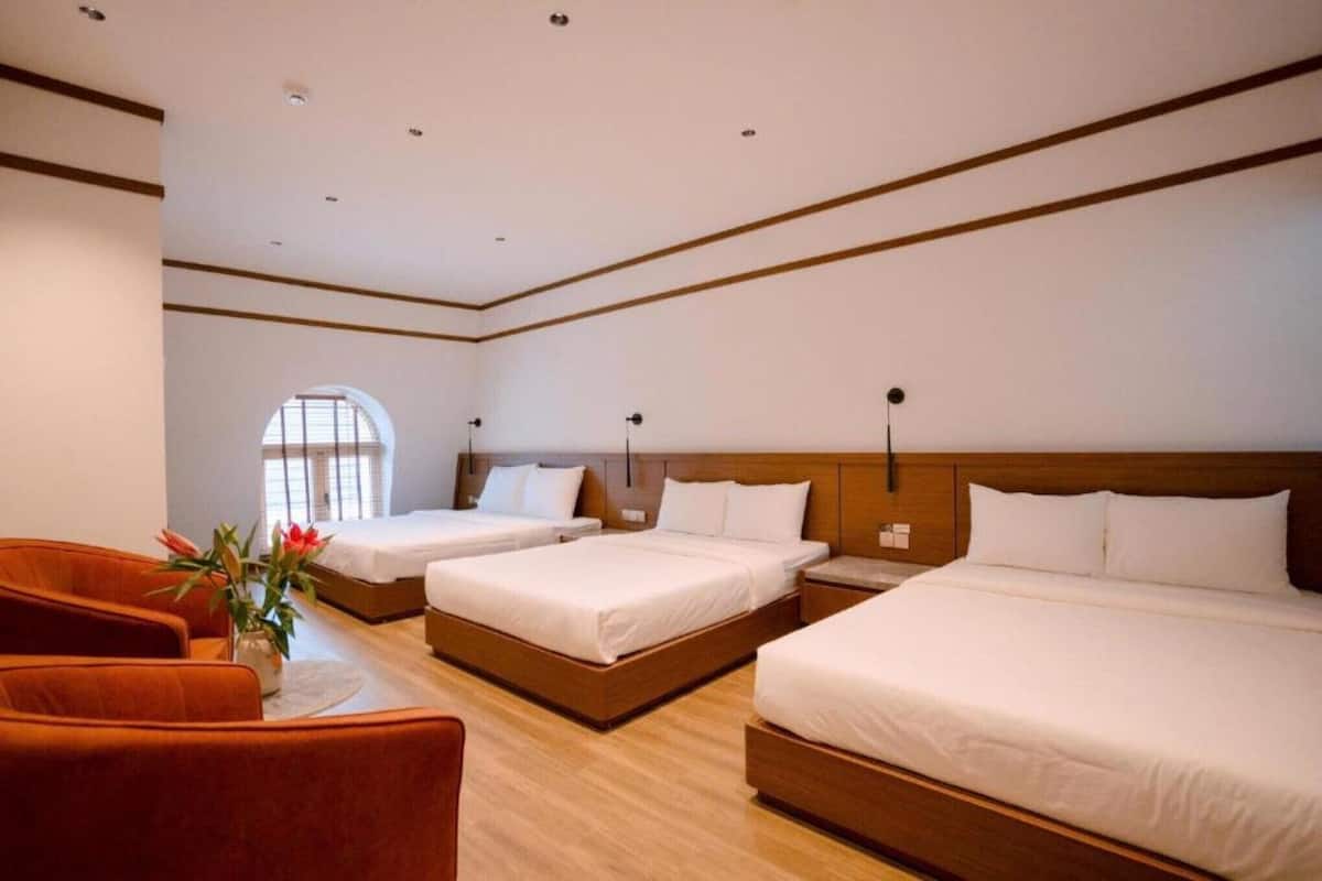 Quarto de hotel com três camas, cabeceira de madeira, janela grande e um vaso com flores.