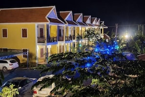 Fila de edifícios amarelos com sacadas, área de estacionamento e árvore com luzes iluminadas.