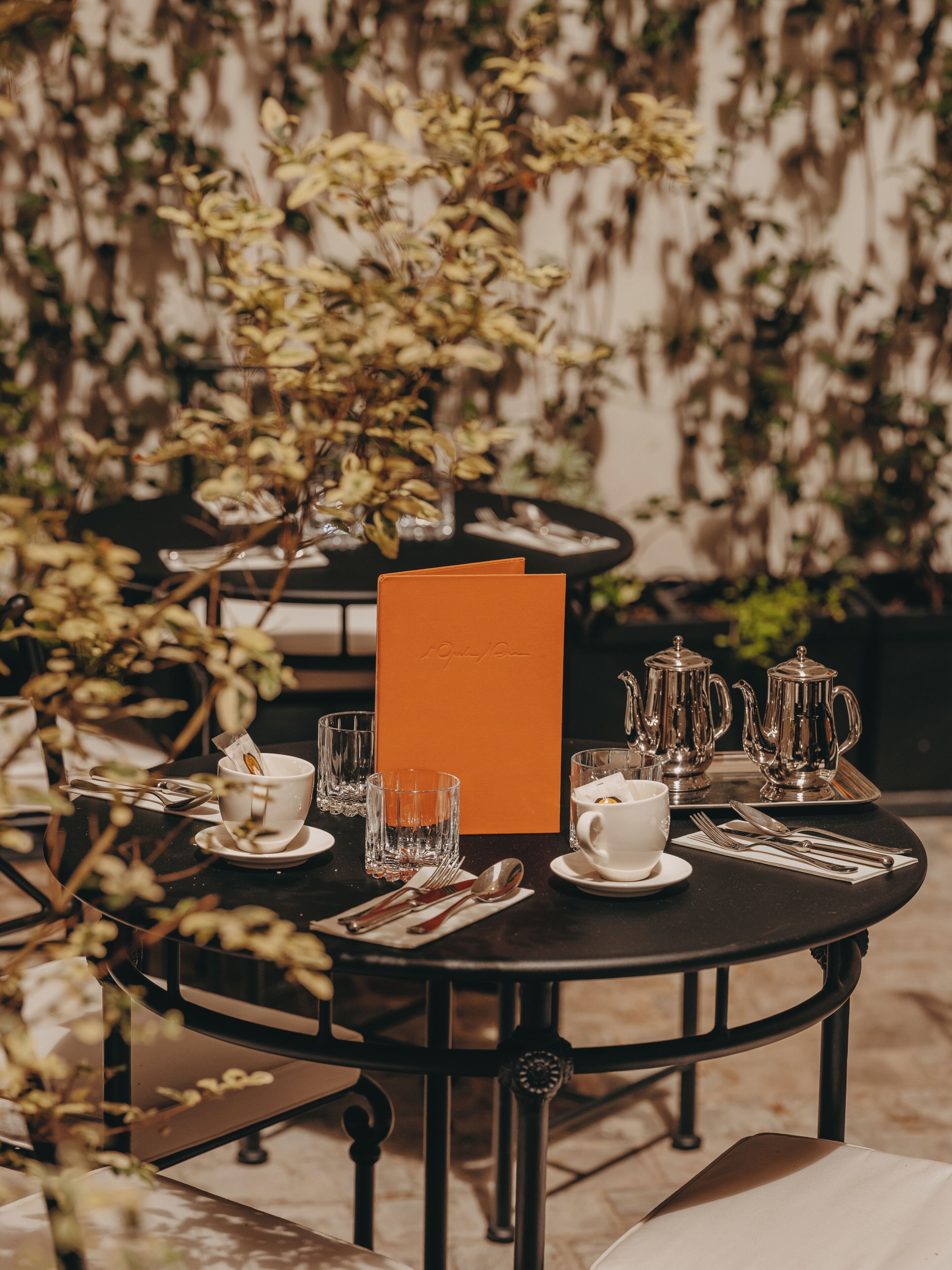 Uma mesa redonda posta com cardápio, bules, xícaras e talheres, rodeada por plantas.