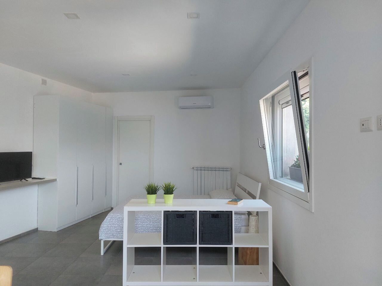 Quarto com parede branca, janela, radiador, armário branco e televisão.
