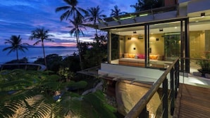 Uma casa moderna com fachada de vidro, deck de madeira e vista para o oceano ao entardecer.