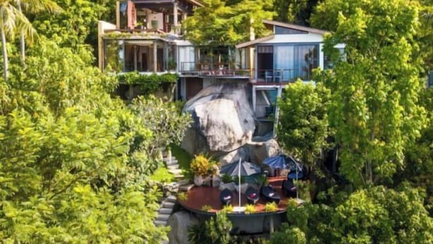 Uma casa de vários andares cercada por vegetação densa, com área de piscina visível e espaço para estar ao ar livre.