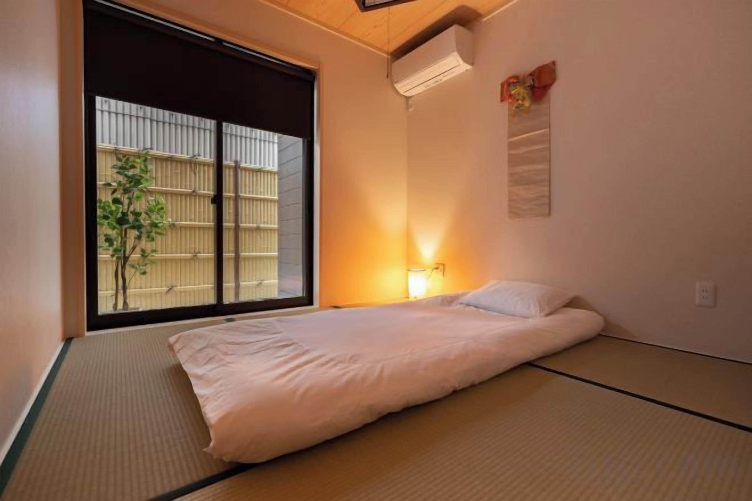 Quarto com piso de tatame, uma cama, janela com vista para uma árvore e ar-condicionado fixado na parede.