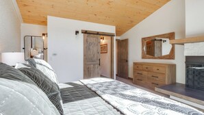 Quarto com teto de madeira, cama com roupa de cama estampada, cômoda, espelho e lareira.