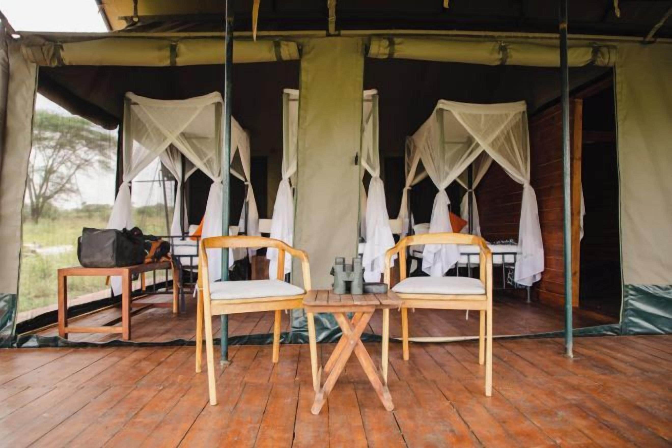 Acomodação em tenda com duas cadeiras de madeira e uma mesa, mosquiteiros e vista para o exterior.