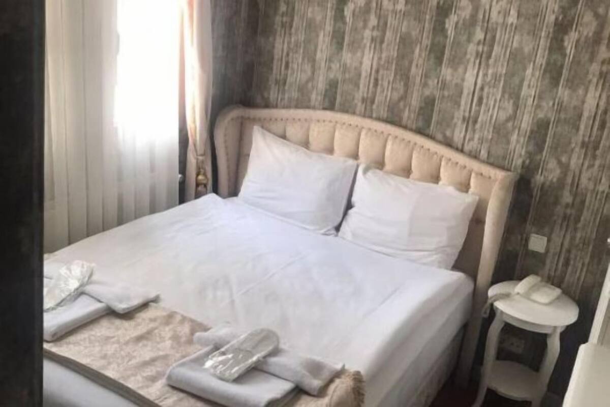 Uma cama bem arrumada com lençóis brancos, uma pequena mesa de cabeceira com um telefone e uma janela com cortinas.