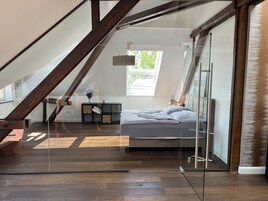 Quarto moderno estilo loft com teto inclinado, porta de correr em vidro, cama com roupa de cama branca e piso de madeira.