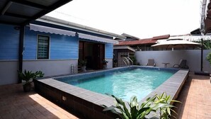 Piscina com borda azulejada, área de varanda coberta e um edifício com revestimento azul e telhado vermelho.