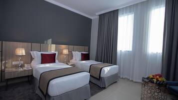 Quarto de hotel com duas camas, uma mesa de cabeceira com abajur, uma mesinha com frutas e uma janela grande com cortinas.