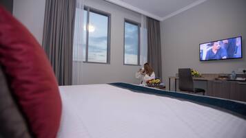 Quarto de hotel com cama, escrivaninha, televisão e janela com cortinas.
