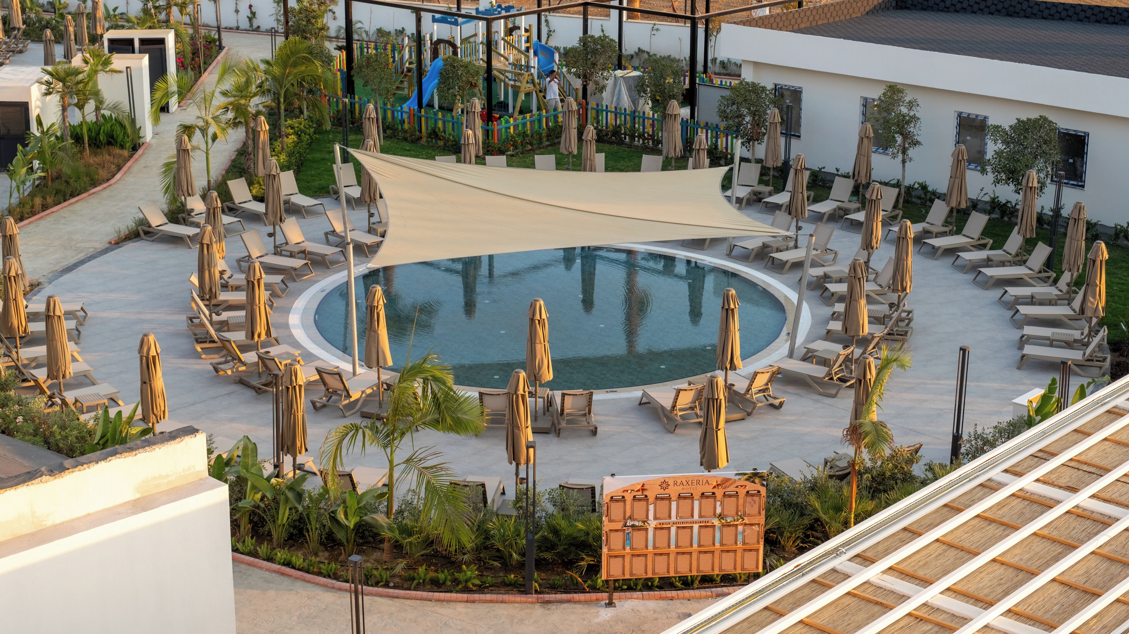 Área de piscina com espreguiçadeiras e guarda-sóis, cercada por vegetação e um parquinho.