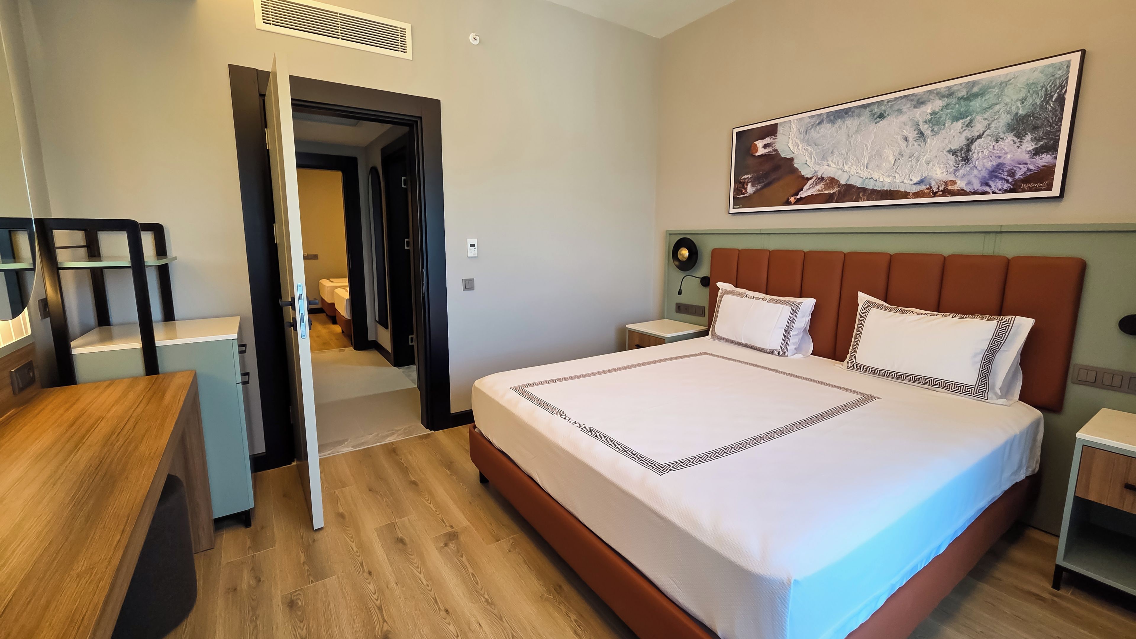 Quarto de hotel com cama, mesas de cabeceira, escrivaninha, porta que dá para outro cômodo e um quadro na parede.