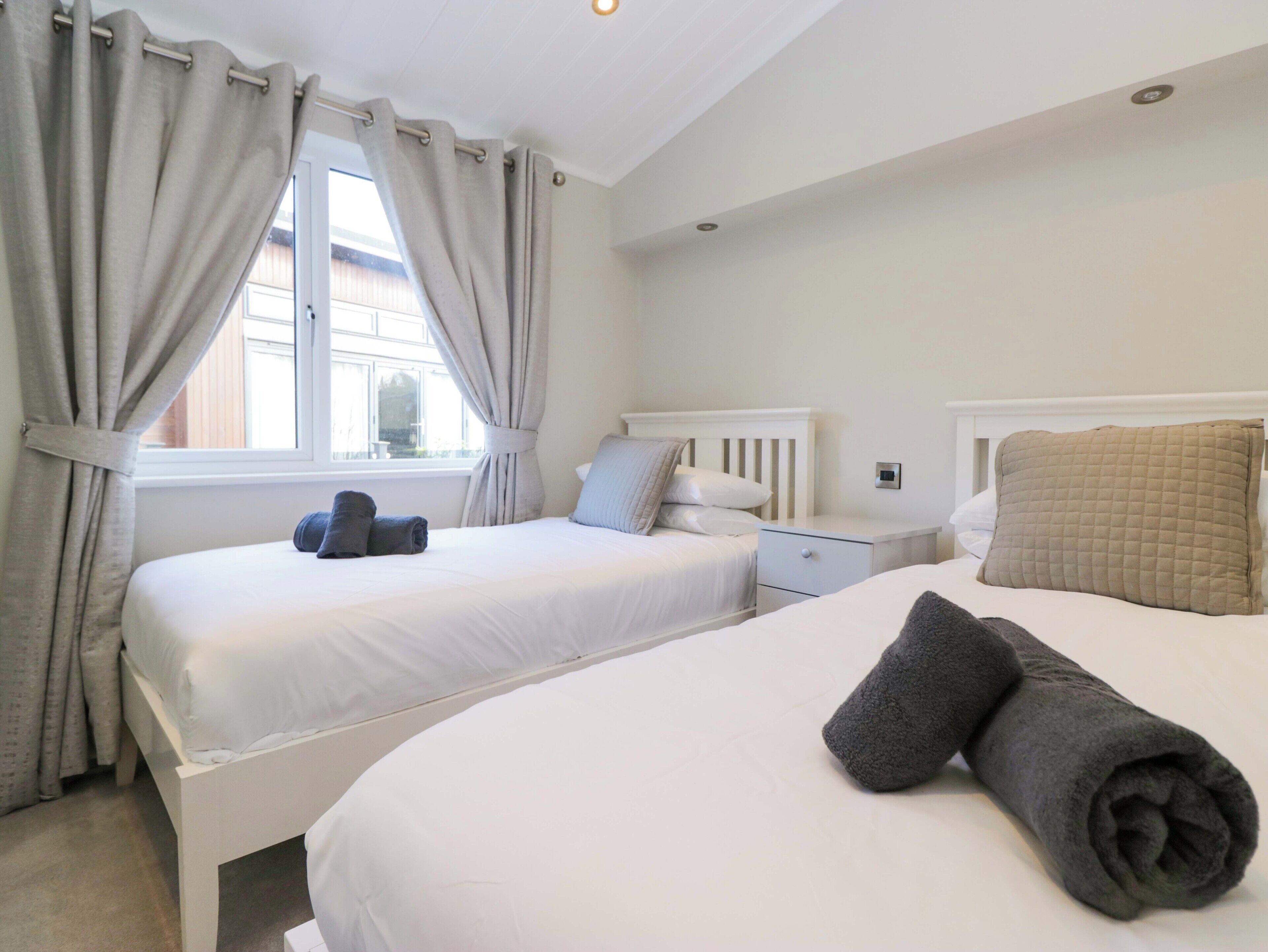 Quarto de hotel com duas camas, janela com cortinas, mesa de cabeceira e peitoris.
