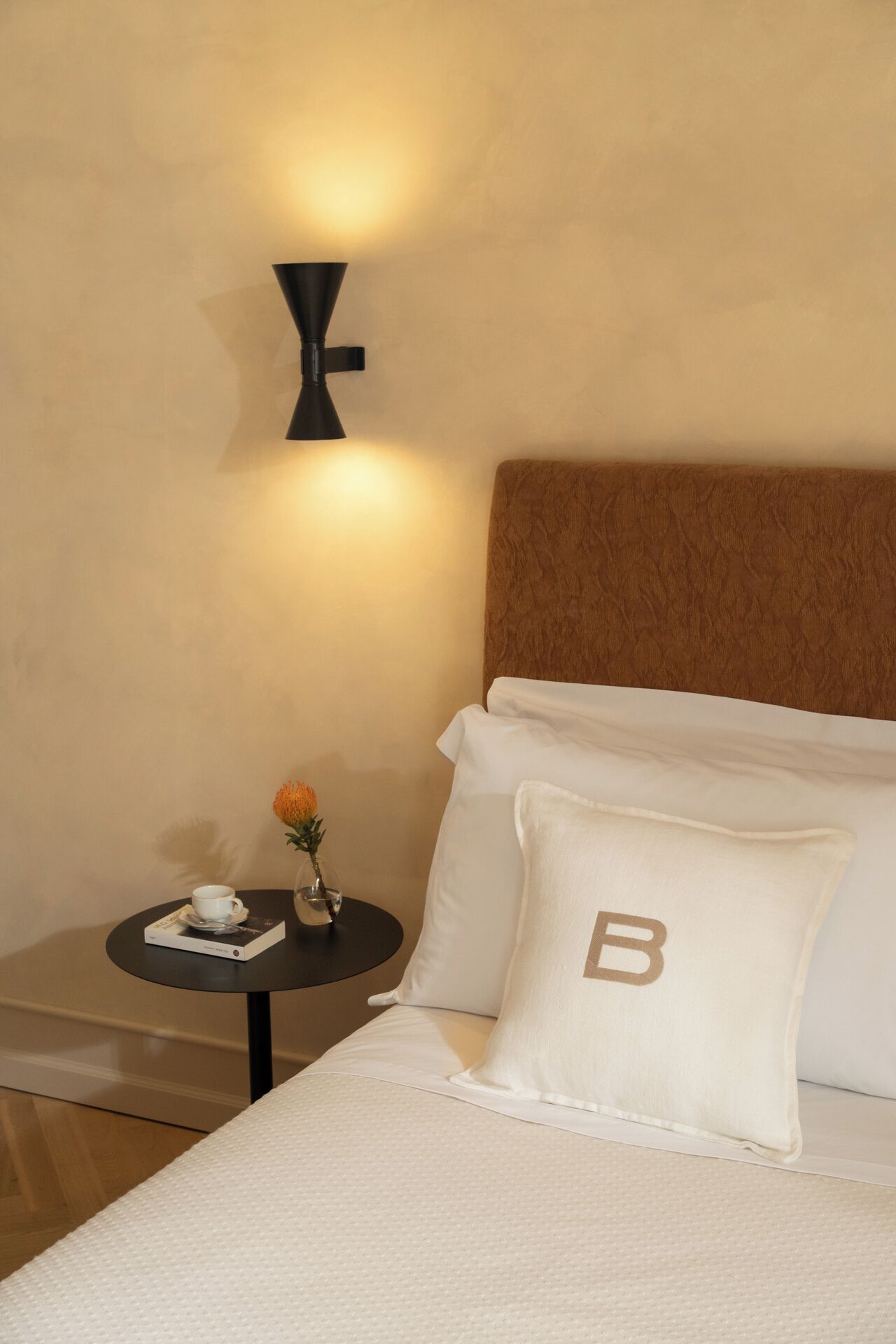 Quarto de hotel com cama bem arrumada, mesa de cabeceira com um livro e um copo, e luminária na parede.