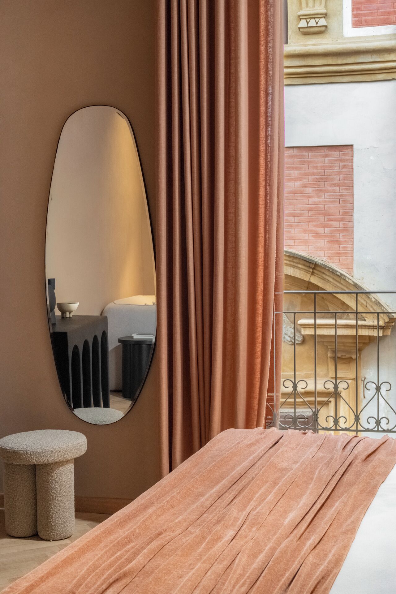 Quarto de hotel com um espelho oval grande, um banco de madeira, uma árvore para gatos em pedra e vista para um pátio com um edifício de tijolos e um portão em ferro forjado.
