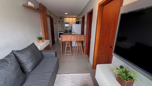 Uma sala de estar moderna com um sofá cinzento, uma mesa de jantar de madeira com dois taburetes, uma cozinha ao fundo e uma televisão grande à direita.