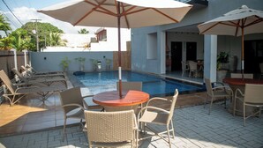 Área de piscina ao ar livre com espreguiçadeiras e guarda-sóis, cercada por um pátio ladrilhado.