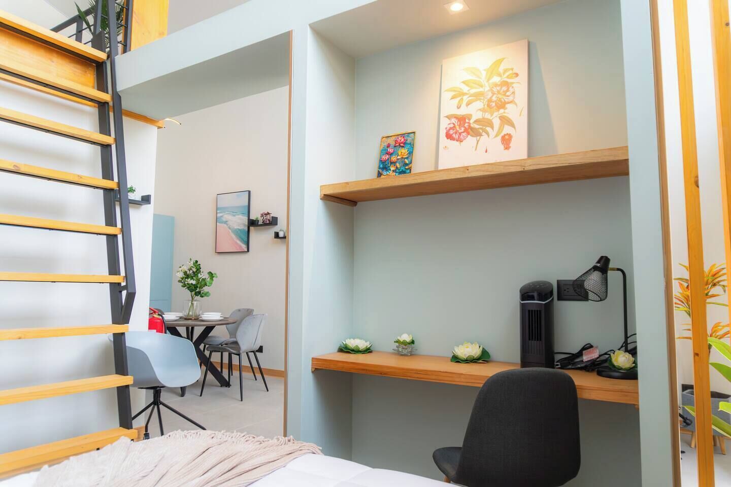 Quarto compacto com cama, escrivaninha, cadeira e uma pequena mesa com uma planta. Há prateleiras com objetos decorativos e um quadro.
