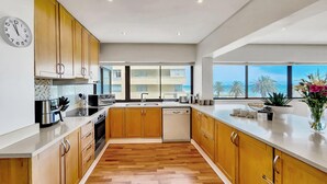 Cozinha moderna com armários de madeira, eletrodomésticos de aço inoxidável e uma janela grande com vista para palmeiras e um edifício.
