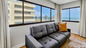 Sala de estar moderna com sofá cinzento, vista para um edifício e para o mar através de grandes janelas.