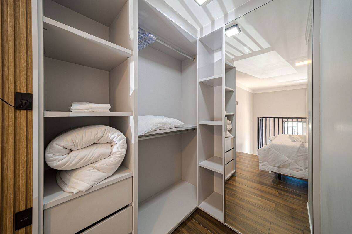 Um closet amplo com prateleiras abertas, uma barra para pendurar roupas e uma cama no quarto vizinho.