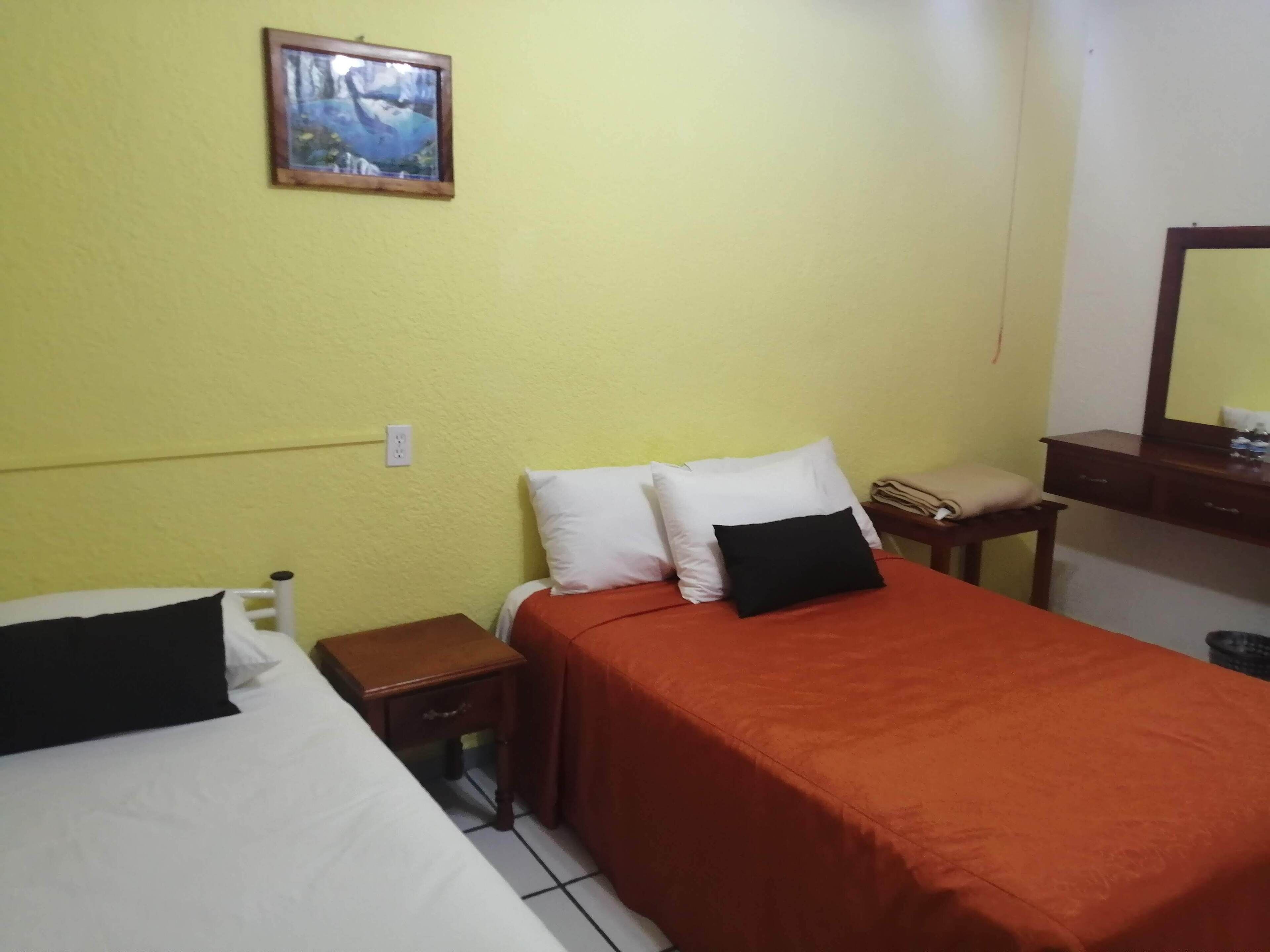 Quarto de hotel com duas camas, uma mesa de cabeceira de madeira, uma escrivaninha com um espelho e um quadro na parede.