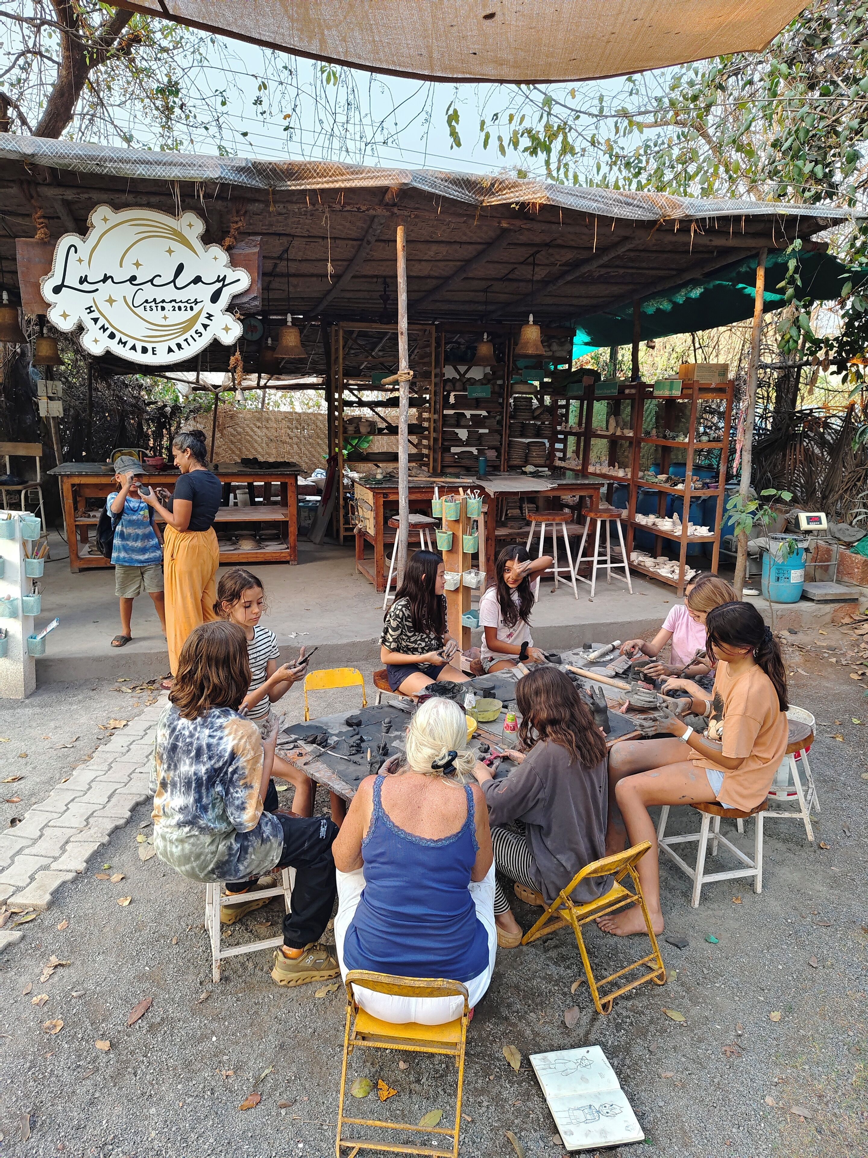 Um grupo de pessoas praticando cerâmica em uma oficina rústica ao ar livre, com uma placa indicando "Luneclay Handmade Artisan".