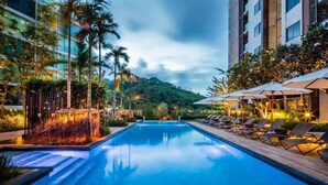 Piscina com espreguiçadeiras e guarda-sóis, cercada por vegetação e edifícios modernos.