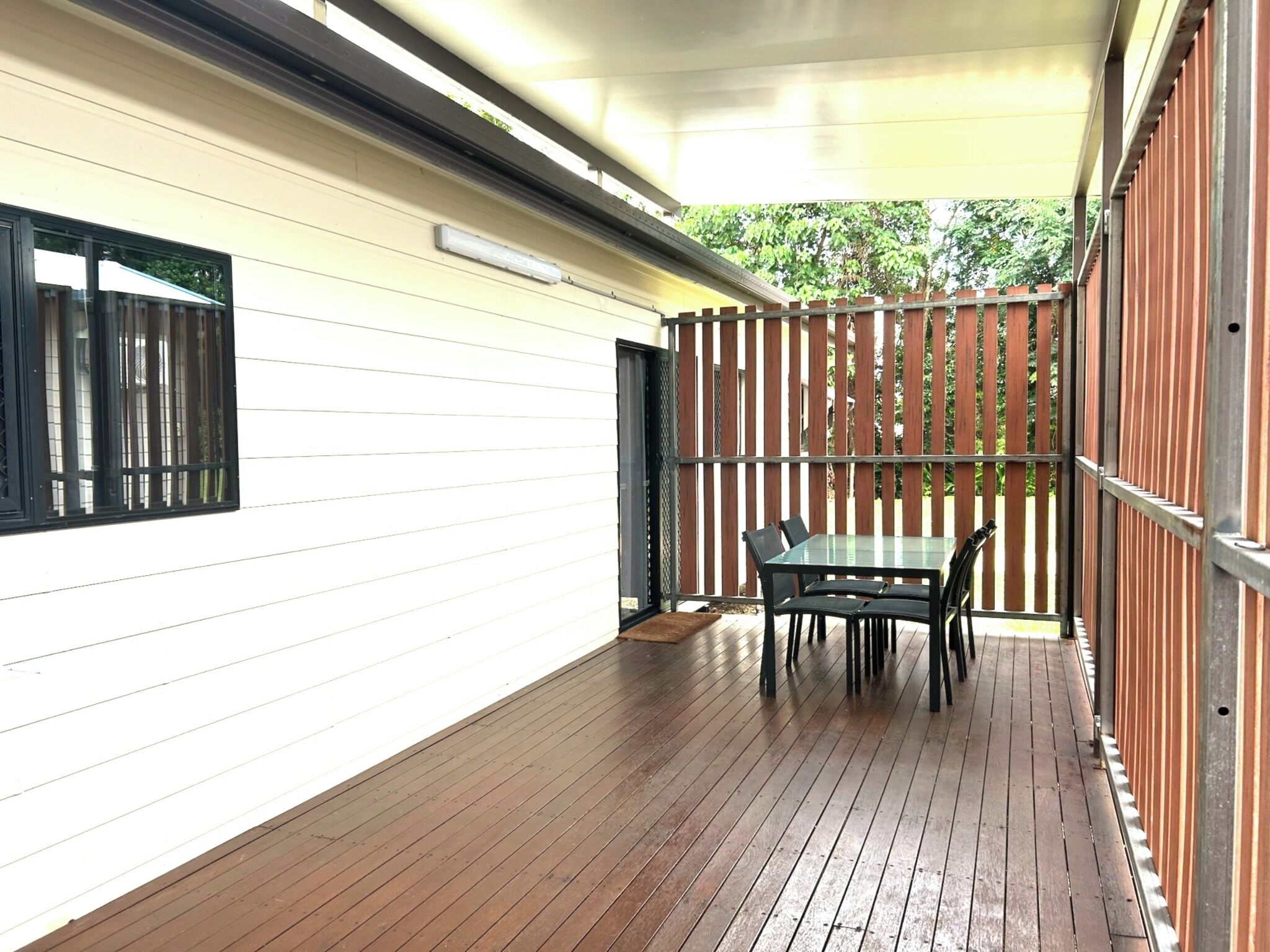 Uma área externa coberta com um deck de madeira, uma mesa com cadeiras e um corrimão de metal.