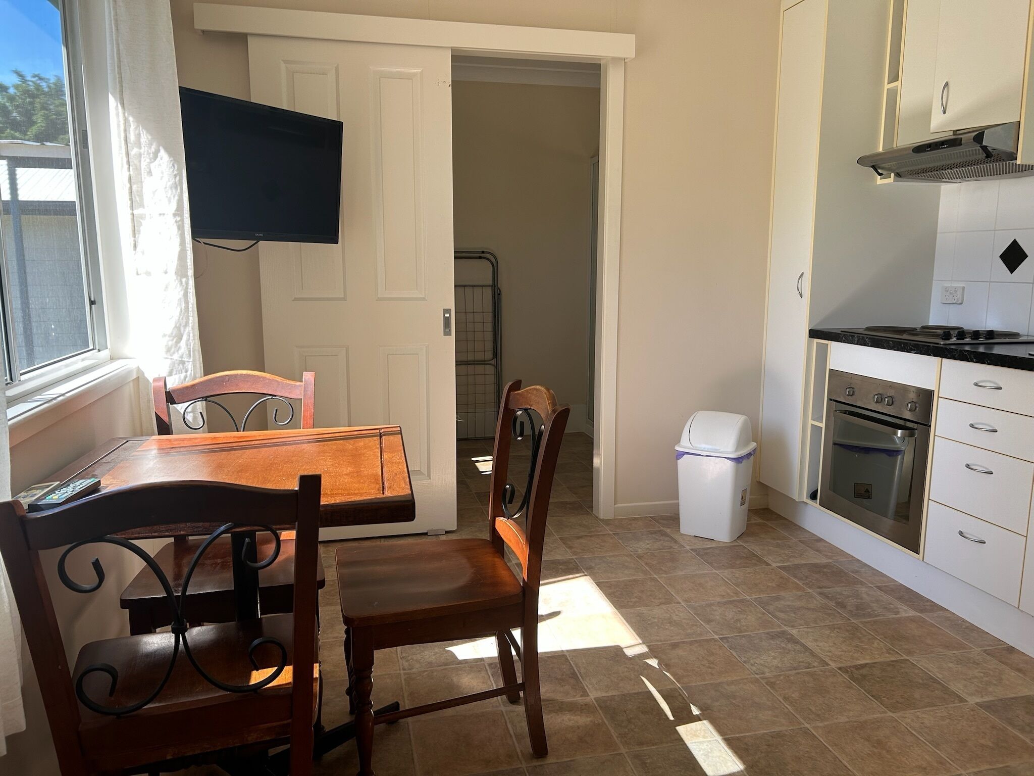 Uma cozinha com uma mesa de jantar, cadeiras, uma TV fixada na parede, uma lixeira e um forno.